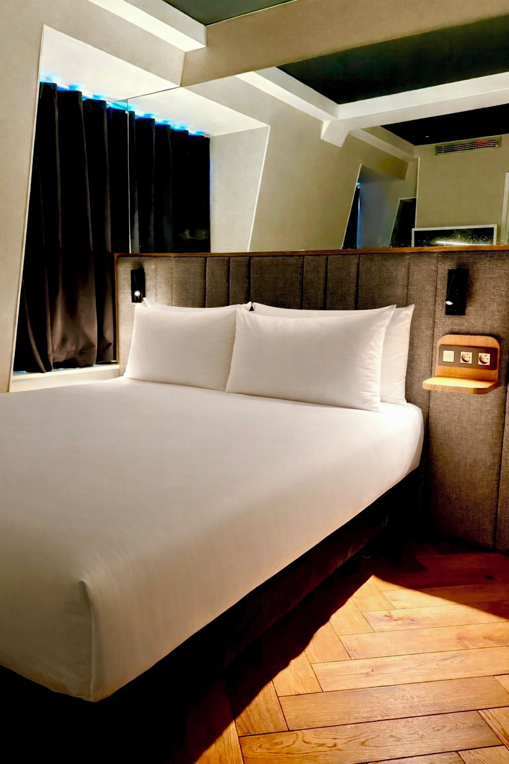 Quarto de hotel moderno com cama grande, piso de madeira e cabeceira embutida com espelho e luminária.