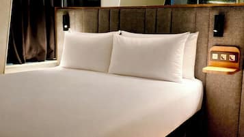 Quarto de hotel moderno com cama grande, piso de madeira e cabeceira embutida com espelho e luminária.