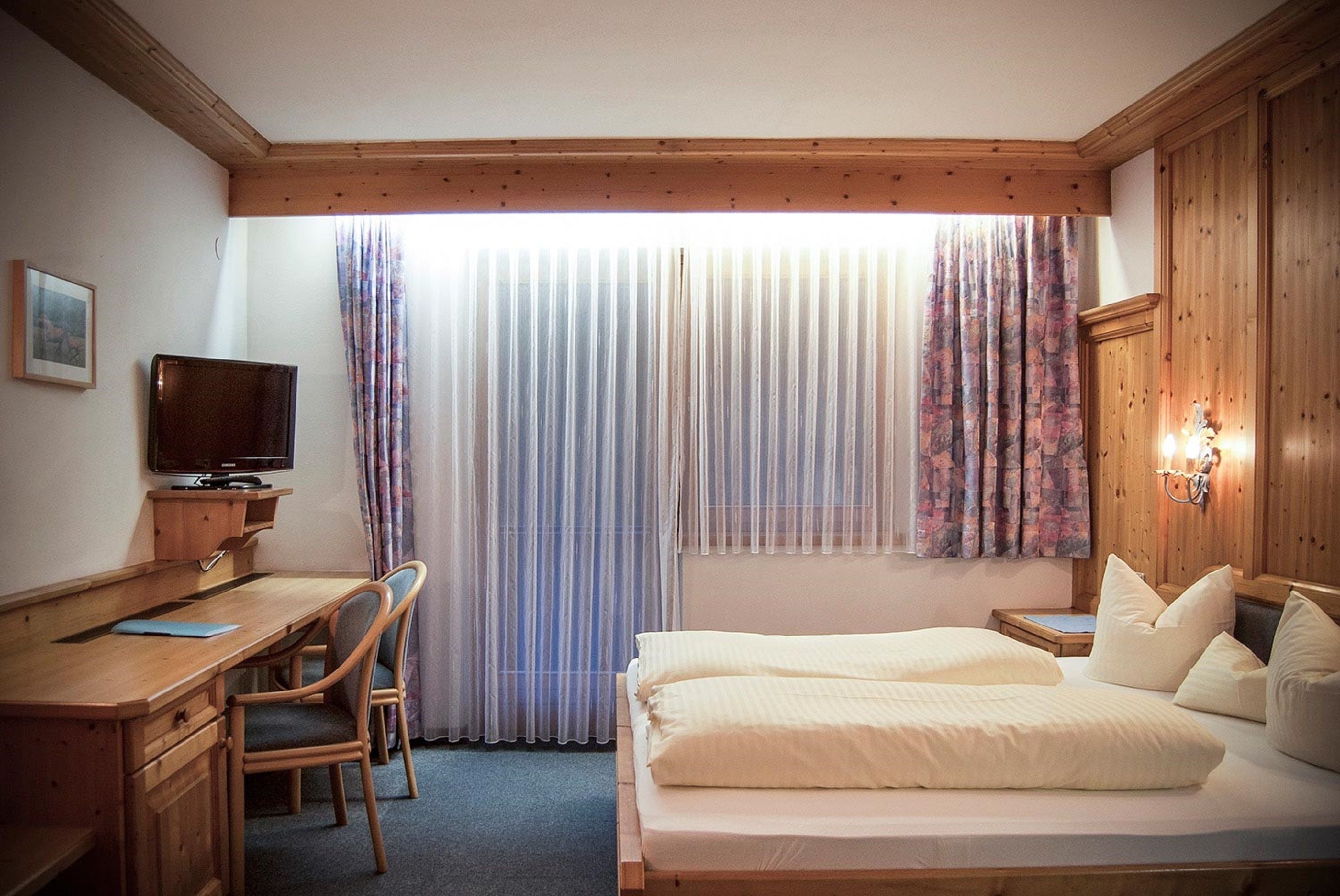 Quarto de hotel com escrivaninha de madeira, duas camas, televisão e janela com cortinas.