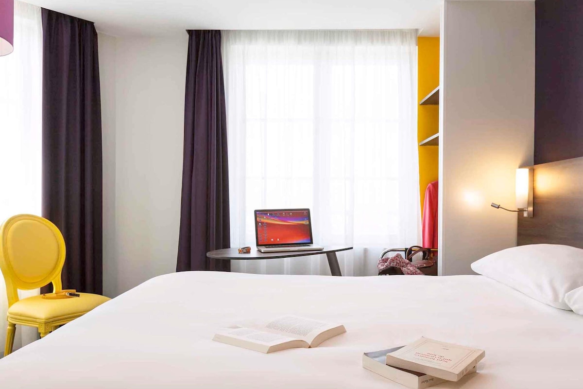 Quarto de hotel com cama, um laptop sobre uma mesinha, uma cadeira e uma mesa de cabeceira com abajur.