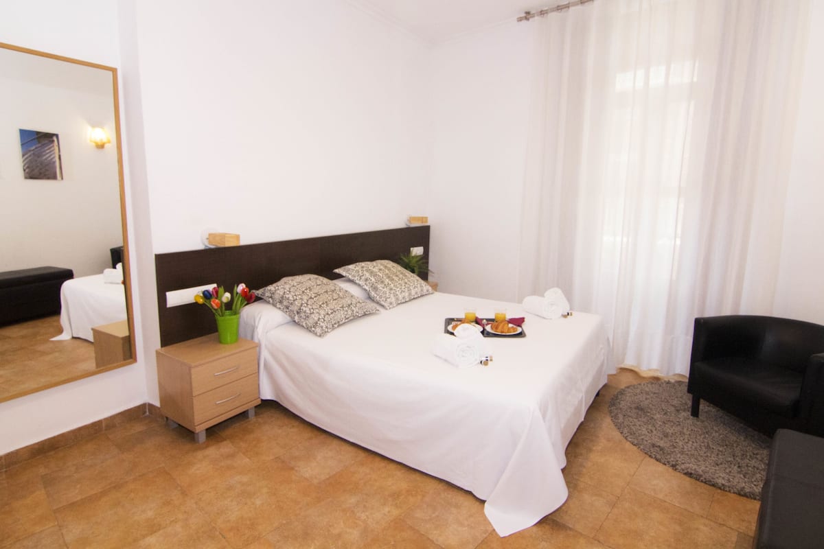 Quarto de hotel com cama, mesa de cabeceira com abajur, cadeira e um espelho grande.