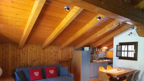 Interior acolhedor de cabana com teto de madeira, sofá azul com almofadas estampadas em formato de coração, área de jantar com mesa e cadeiras de madeira e cozinha com janela.