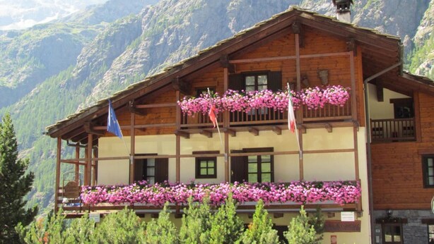 Uma chalé de madeira com varandas decoradas com flores cor-de-rosa, ao fundo montanhas.