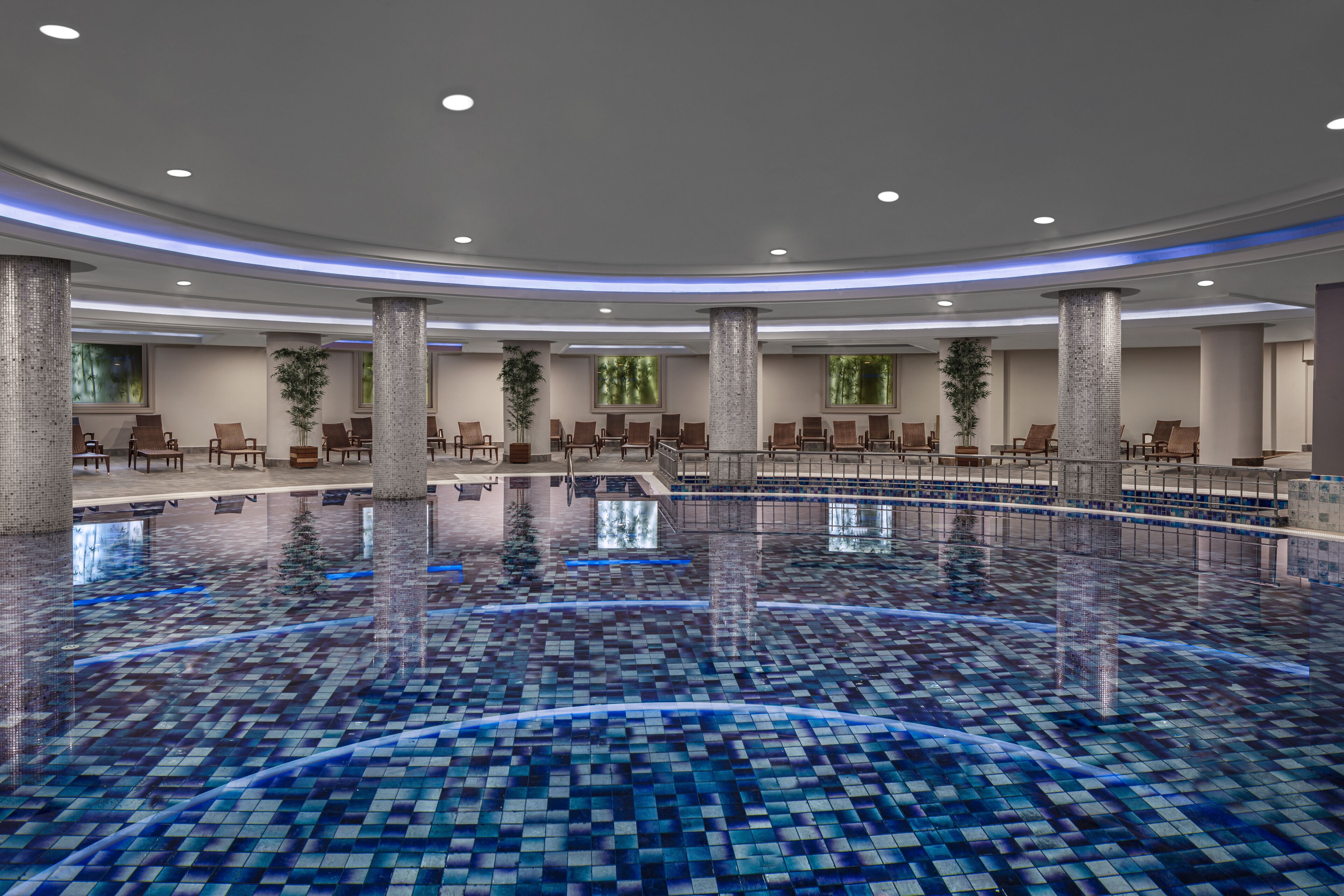 Uma ampla área de piscina interna com piso de azulejo, vários espaços para sentar e grandes pilares.