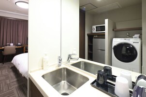 Quarto de hotel com cama, escrivaninha, pia, espelho, máquina de lavar e micro-ondas.