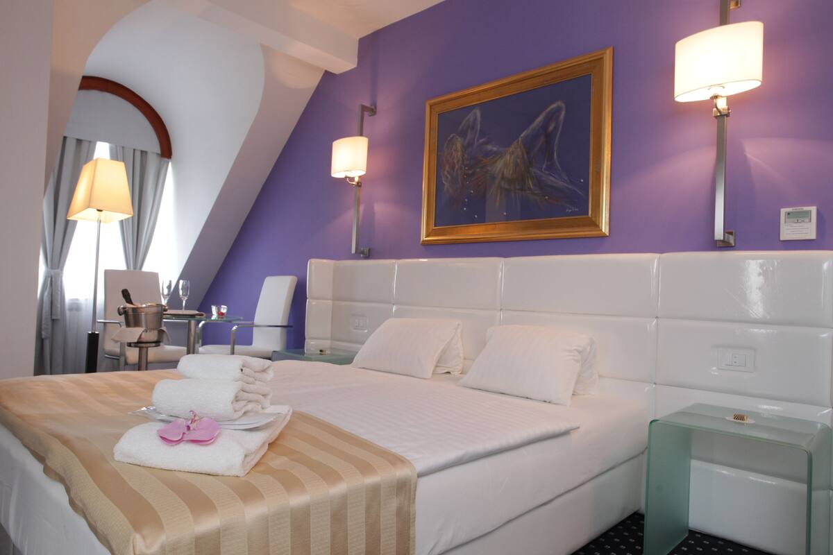 Quarto de hotel com cama, duas luminárias de cabeceira, um quadro na parede, uma mesinha com abajur e uma janela com cortinas.