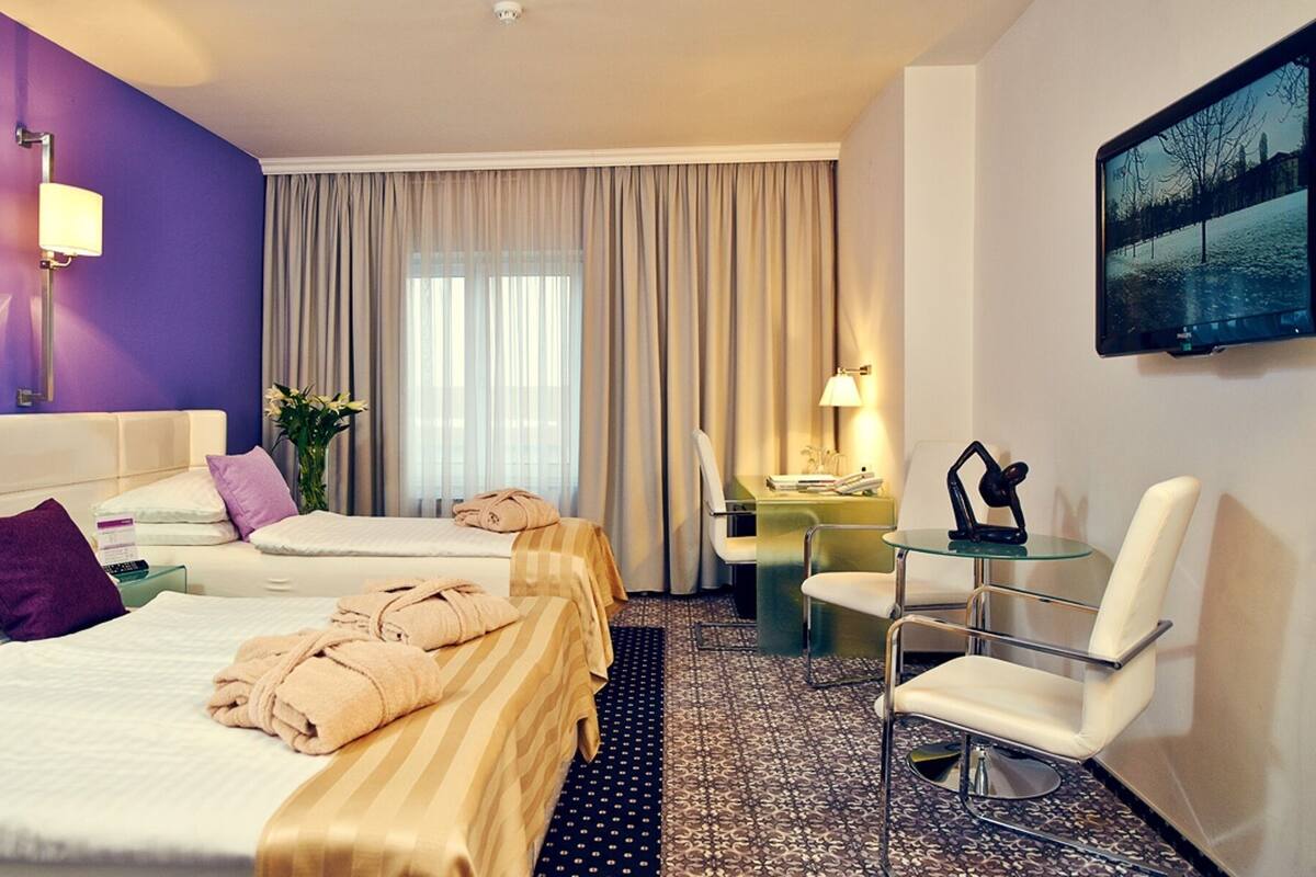 Quarto de hotel com duas camas, uma escrivaninha, uma cadeira e uma televisão na parede.