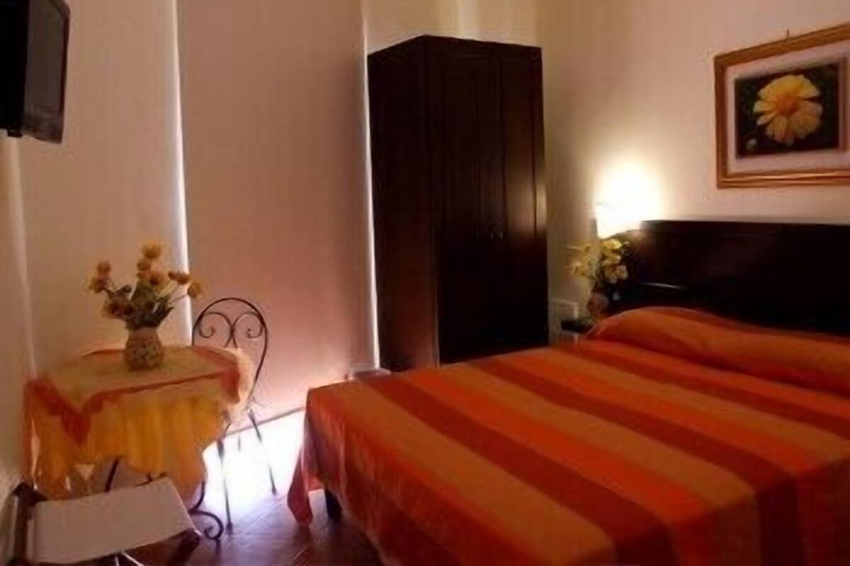 Quarto de hotel com cama, uma mesinha com um vaso de flores, uma cadeira e um guarda-roupa escuro.