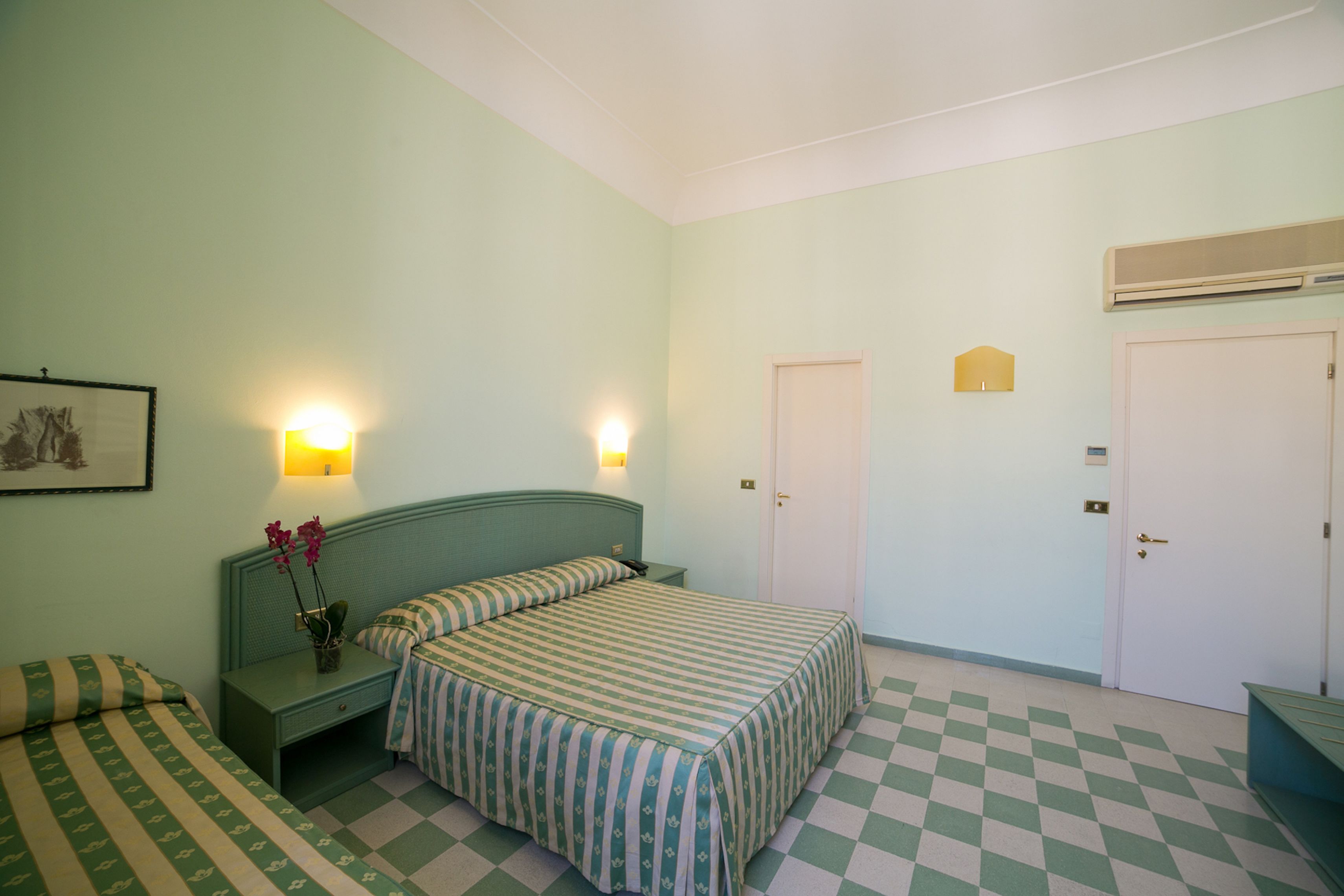 Quarto com piso xadrez, cama com roupa de cama listrada, duas mesas de cabeceira com luminárias e um ar-condicionado fixado na parede.