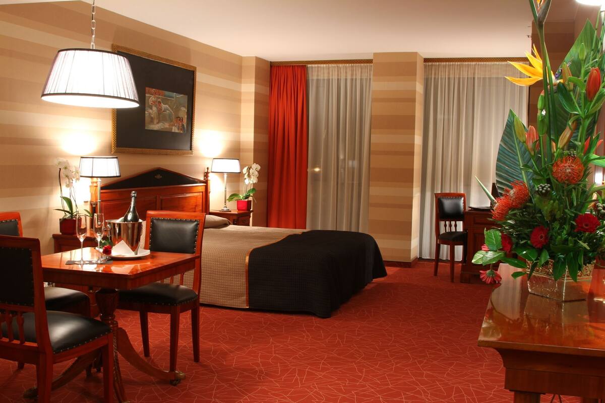 Quarto de hotel com cama, escrivaninha, cadeira, luminária, quadro na parede e planta.