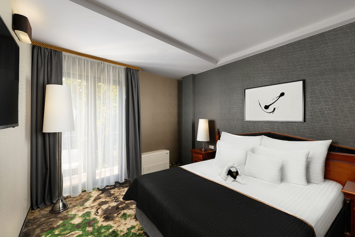 Quarto de hotel com uma cama grande, duas mesas de cabeceira, uma televisão de tela plana, uma janela com cortinas translúcidas e uma obra de arte emoldurada na parede.