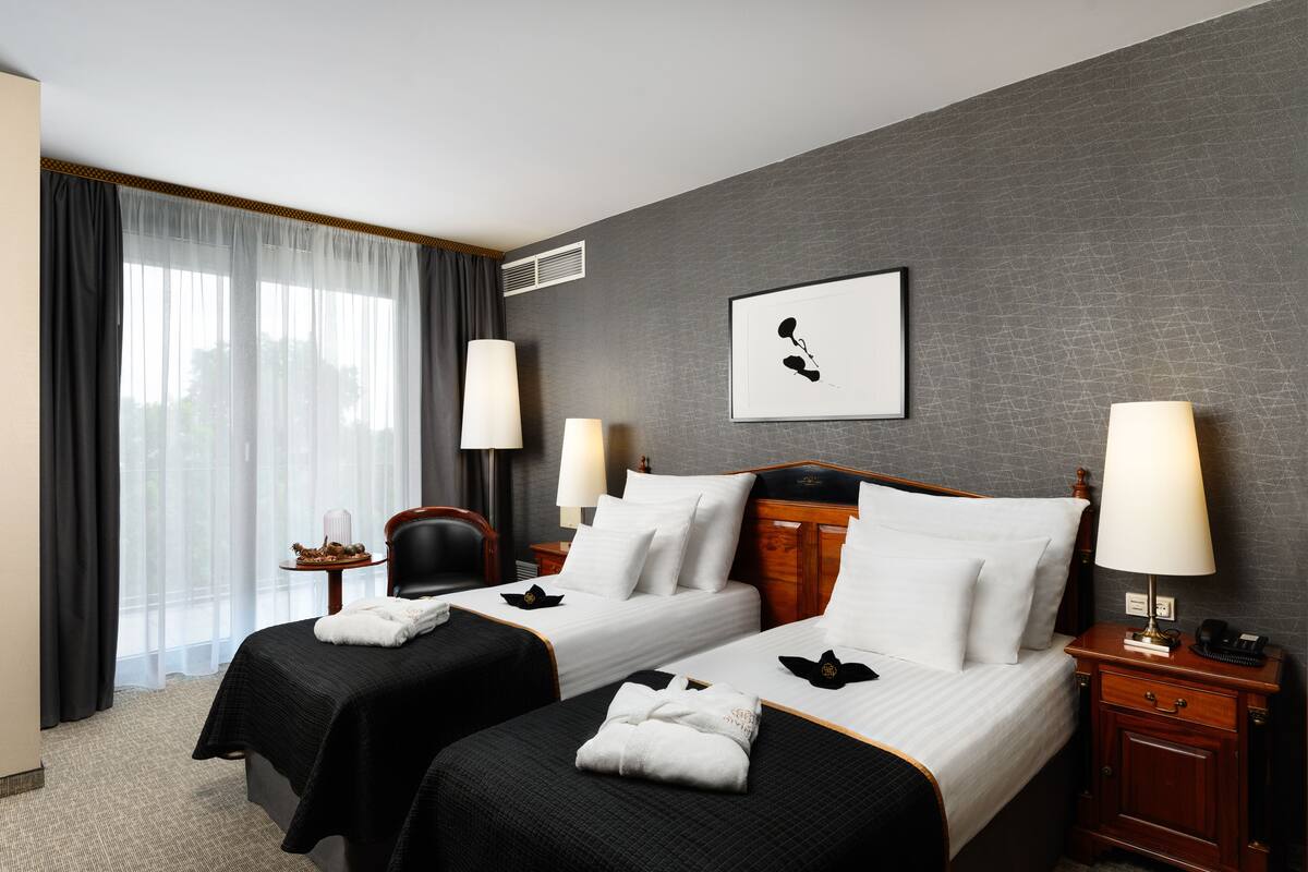 Quarto de hotel com duas camas, uma janela grande com cortinas, um quadro na parede e uma mesa de cabeceira com abajur.