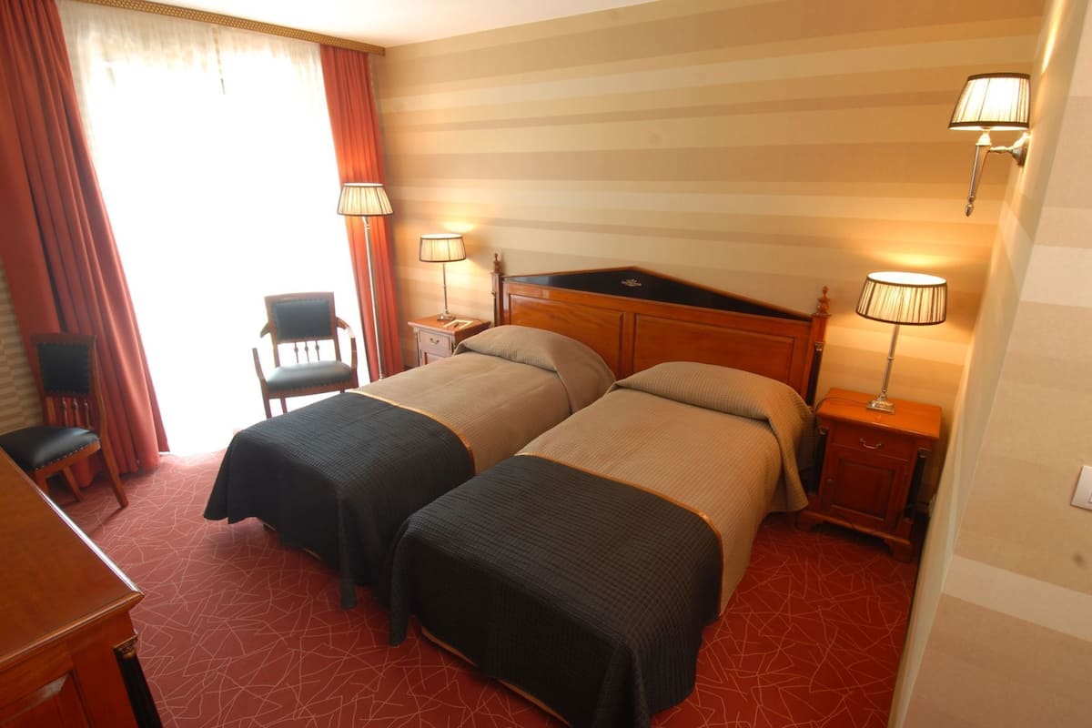 Quarto de hotel com duas camas, uma escrivaninha, uma cadeira e duas luminárias de cabeceira.