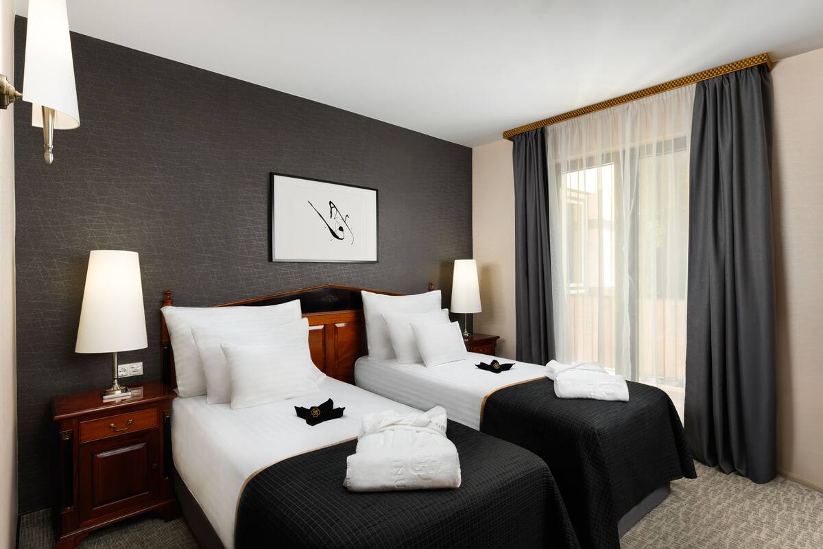 Quarto de hotel com duas camas, parede escura com textura, uma obra de arte emoldurada e uma janela com cortinas.