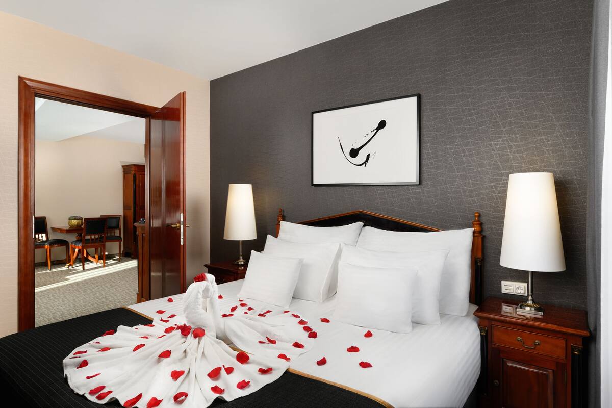 Quarto de hotel com uma cama grande, duas mesas de cabeceira, um painel de madeira na cabeceira e uma obra de arte emoldurada na parede, além de vista para o quarto vizinho.