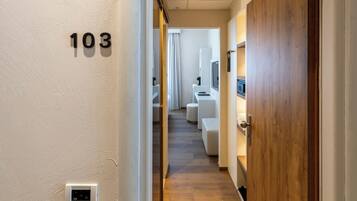 Um corredor com o número '103' na parede, que leva a um quarto com porta de madeira e parede branca.