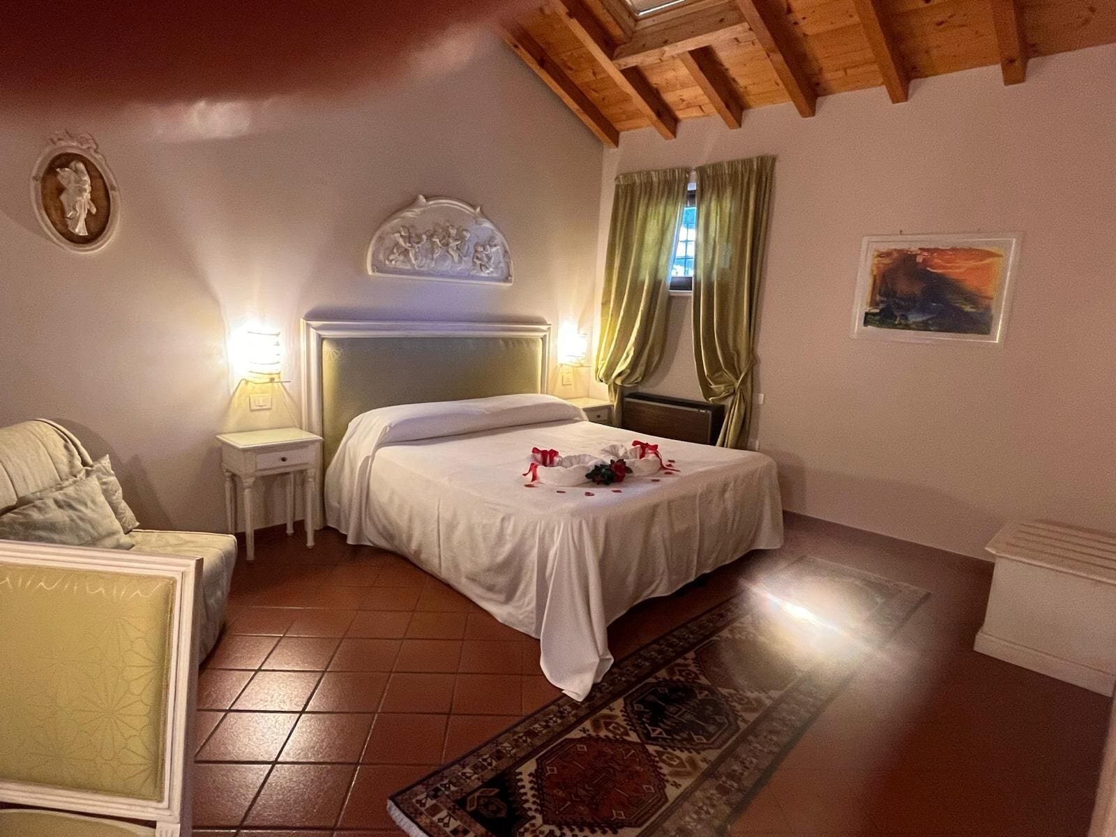 Quarto de hotel com cama, poltrona, mesa de cabeceira, abajur, janela com cortinas e um quadro na parede.