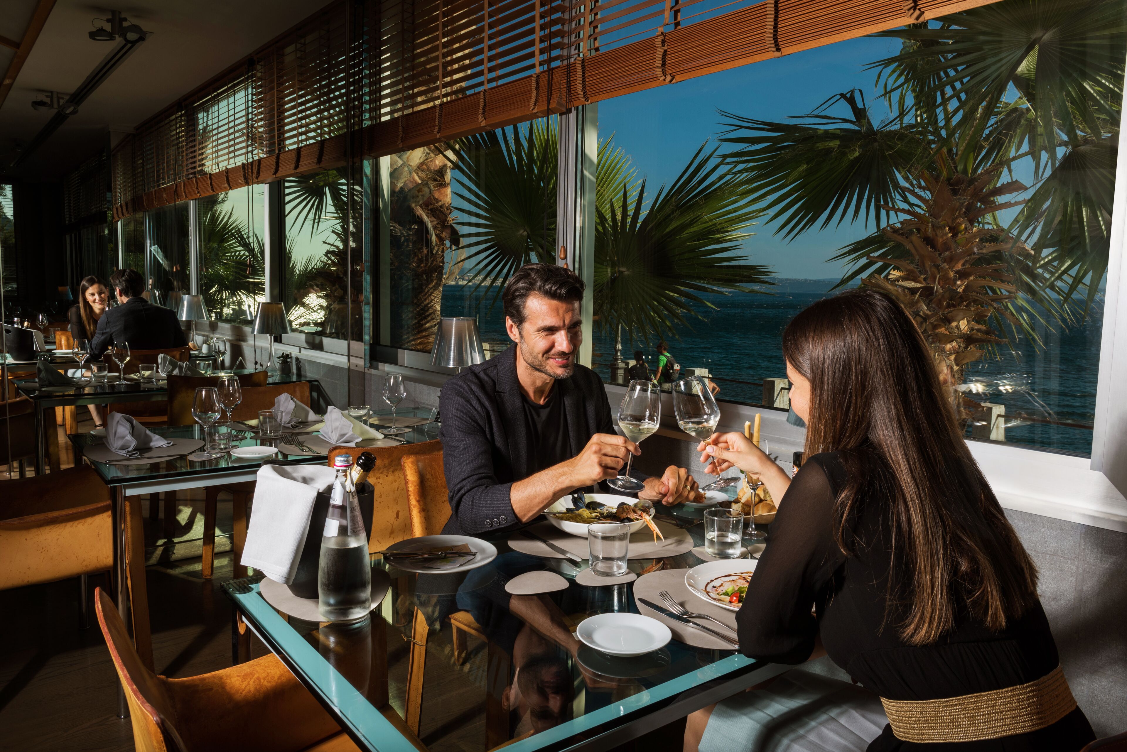 Um restaurante com vista para o mar, com mesas de vidro, cadeiras e utensílios de jantar como pratos, copos e talheres.