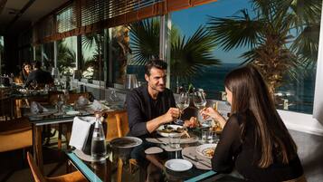 Um restaurante com vista para o mar, com mesas de vidro, cadeiras e utensílios de jantar como pratos, copos e talheres.