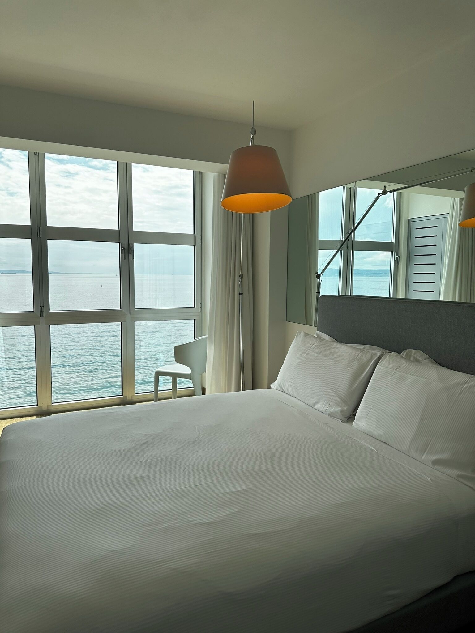 Uma cama grande com roupa de cama branca, cabeceira cinza e vista para o mar através de janelas do chão ao teto.