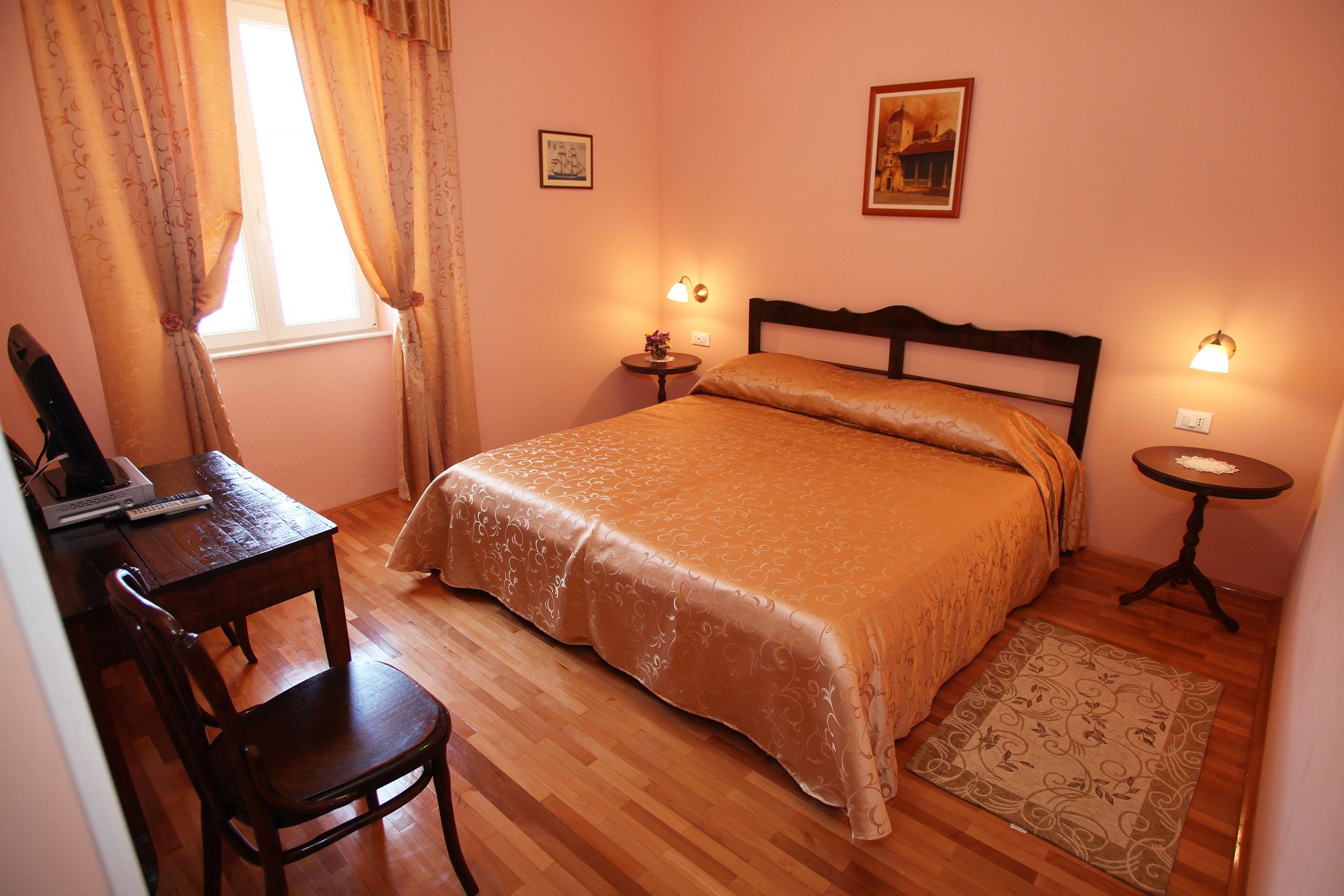 Quarto com cama, escrivaninha com computador, duas luminárias de cabeceira, uma cadeira e um tapete.