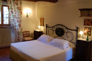 Cama bem arrumada com lençóis brancos, cabeceira de madeira, mesa de cabeceira com abajur, uma cadeira e um relógio fixado na parede.