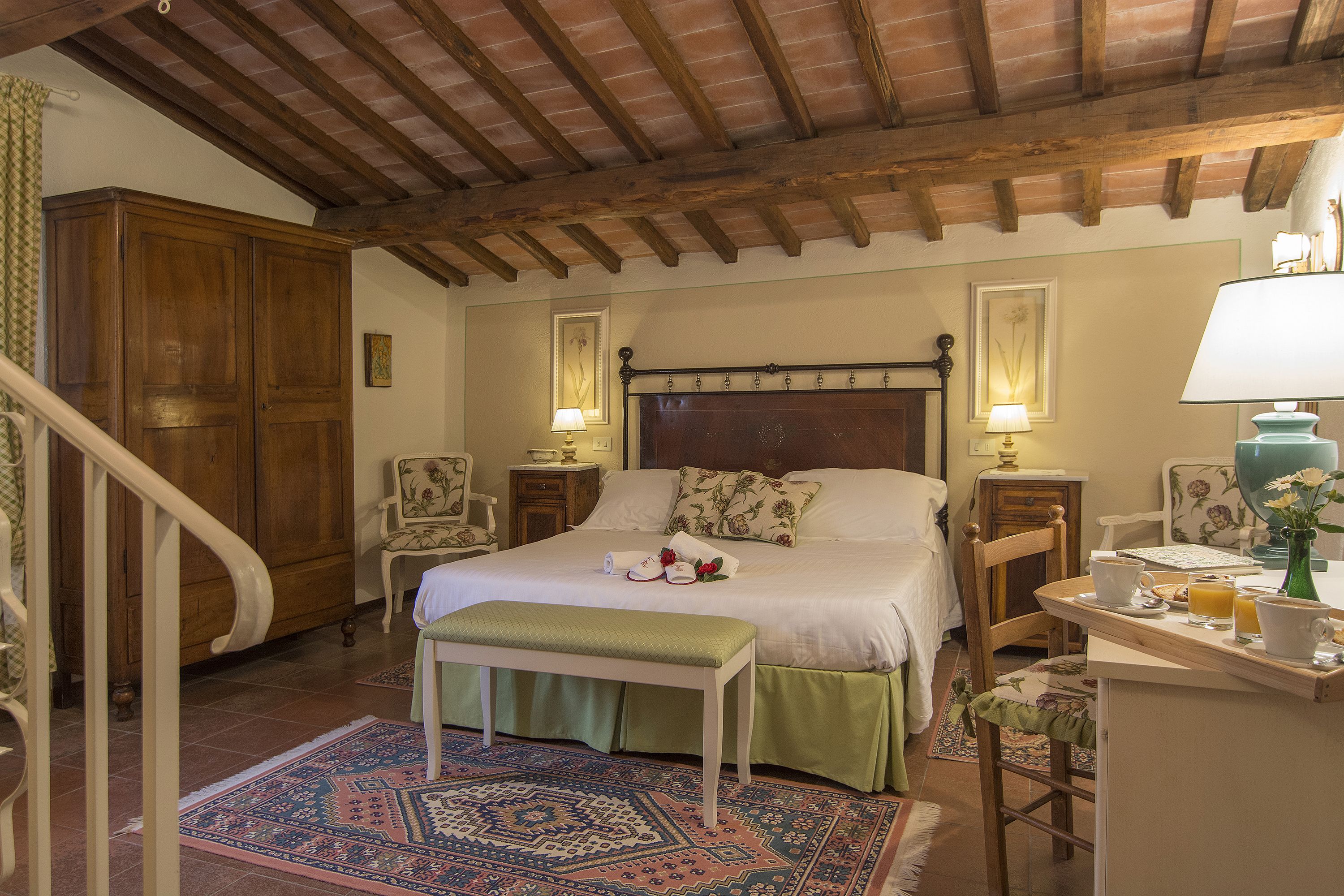 Quarto com teto de madeira, uma cama grande com almofadas florais, uma mesinha de cabeceira com abajur e uma cadeira com almofada estampada.