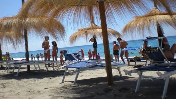 Uma praia com guarda-sóis de palha, espreguiçadeiras e pessoas aproveitando o mar.