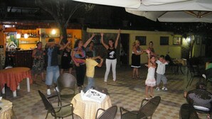 Uma área de refeições ao ar livre com mesas e cadeiras, um bar ao fundo e pessoas dançando.