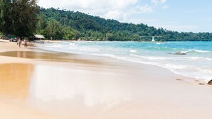 Uma praia com areal, uma colina arborizada ao fundo e um farol à distância.