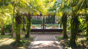 Um caminho no jardim com um banco e uma mesa, cercado por vegetação exuberante e palmeiras.