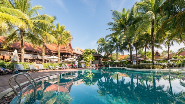Uma piscina com espreguiçadeiras e guarda-sóis, cercada por palmeiras e edifícios com telhados vermelhos.