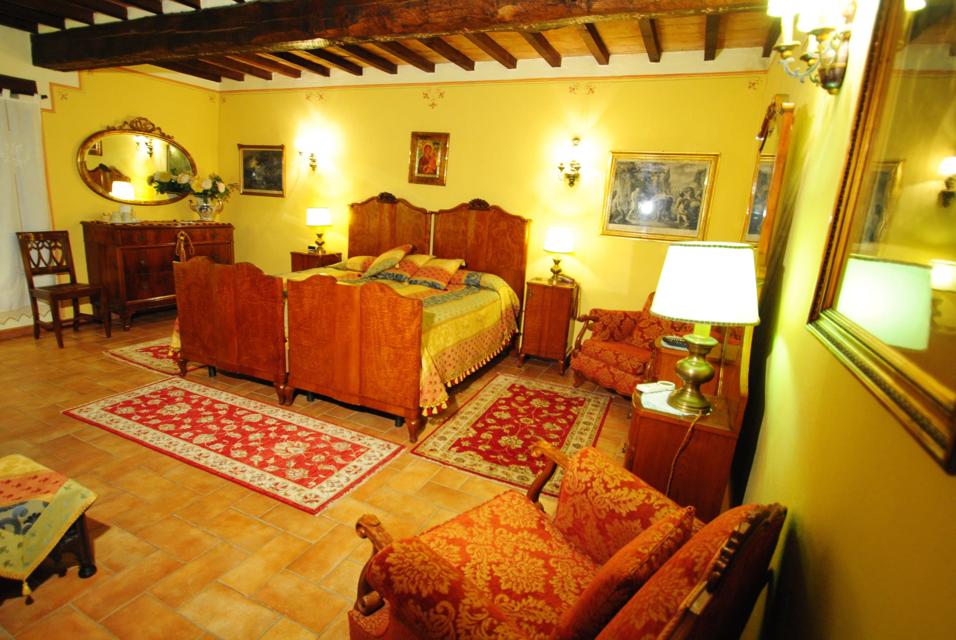 Quarto tradicional com cama de madeira, duas mesas de cabeceira com abajures, uma cadeira e uma pequena mesa com um vaso.