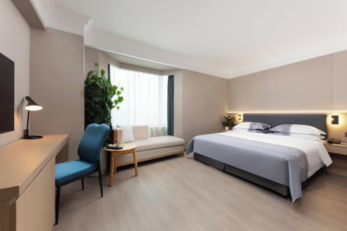 Quarto de hotel moderno com cama, mesa de cabeceira, cadeira, escrivaninha e uma janela grande com cortinas.