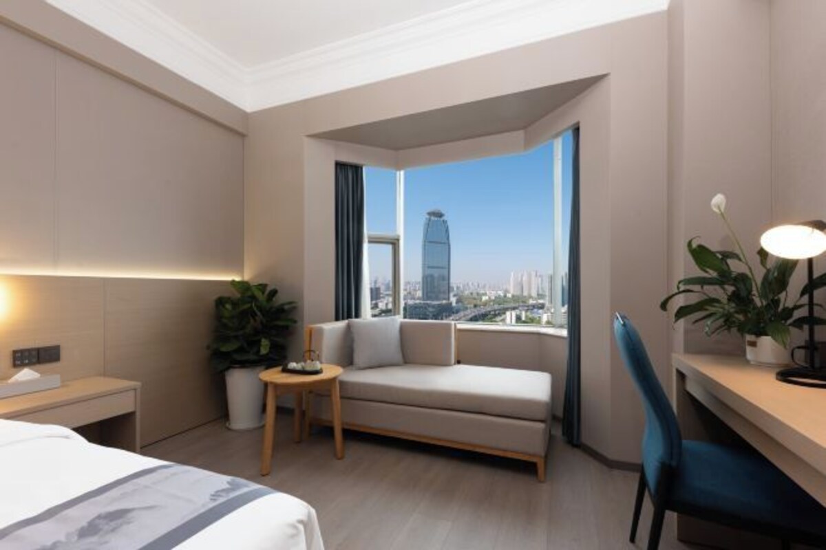 Quarto de hotel com cama, sofá, escrivaninha, cadeira, mesa pequena, planta em vaso e uma grande janela com vista para a cidade.
