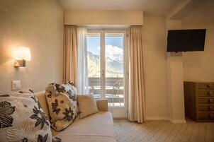 Quarto de hotel com sofá, televisão de tela plana, janela com cortinas e vista para montanhas.