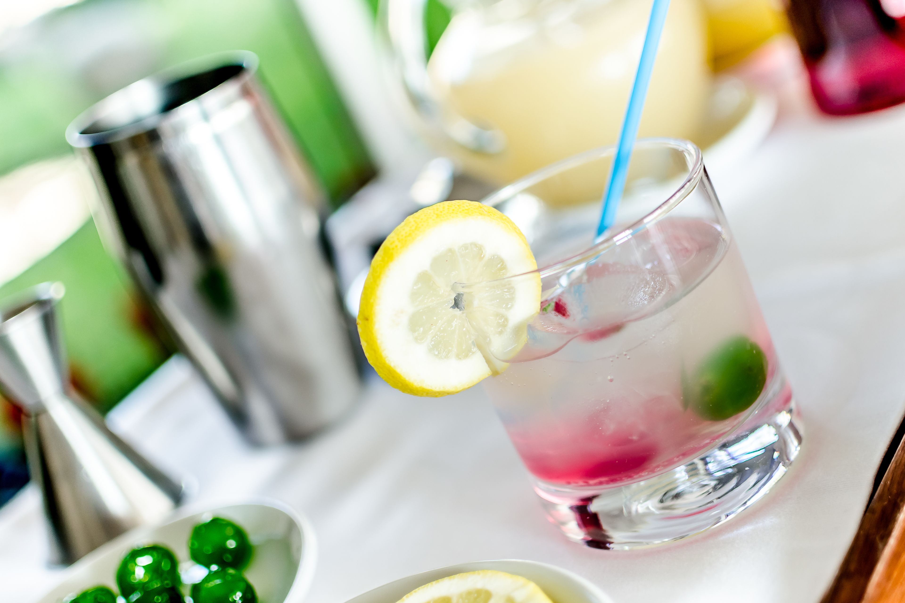 Um copo de cocktail com uma fatia de limão e um canudo azul, rodeado por shaker de cocktail e um jarro.