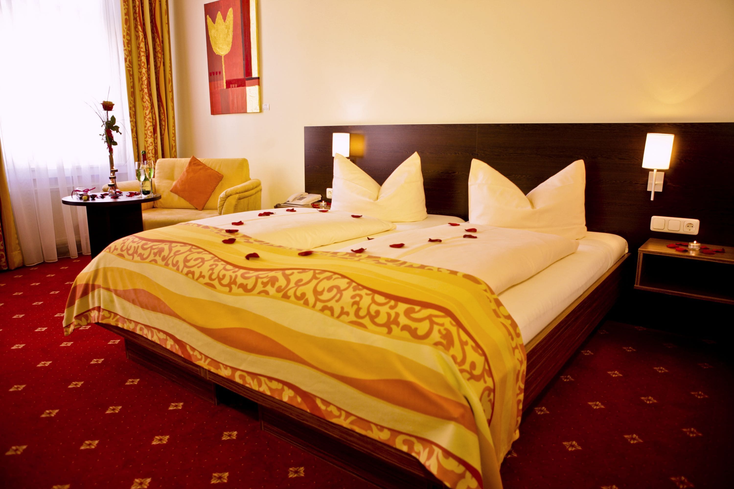 Quarto de hotel com uma cama grande, uma mesinha de cabeceira, uma cadeira, uma pequena mesa com um vaso, e uma parede com um quadro.