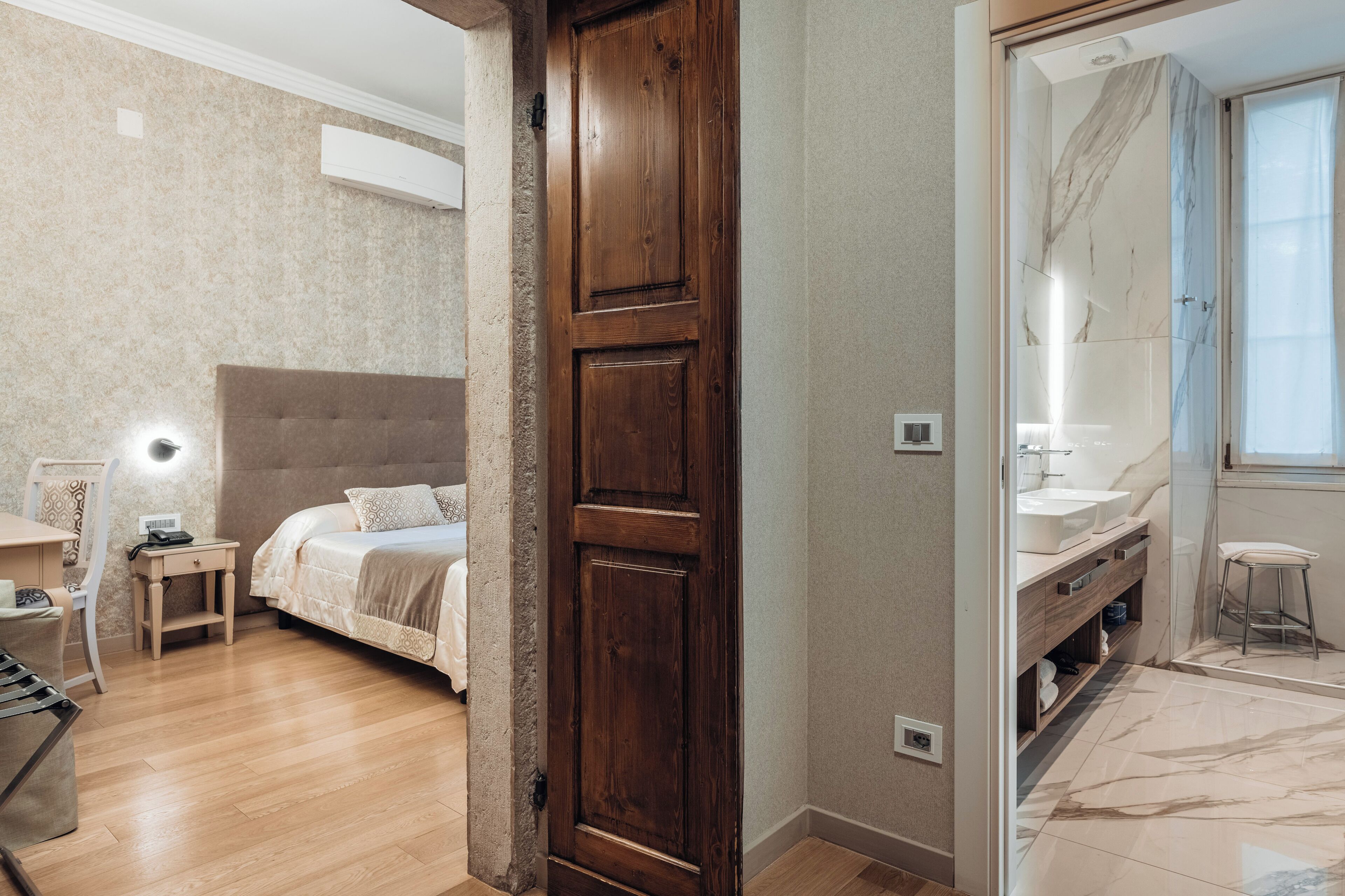 Um quarto com porta de madeira, uma cama com cabeceira, mesa de cabeceira e apoio para luminária.
