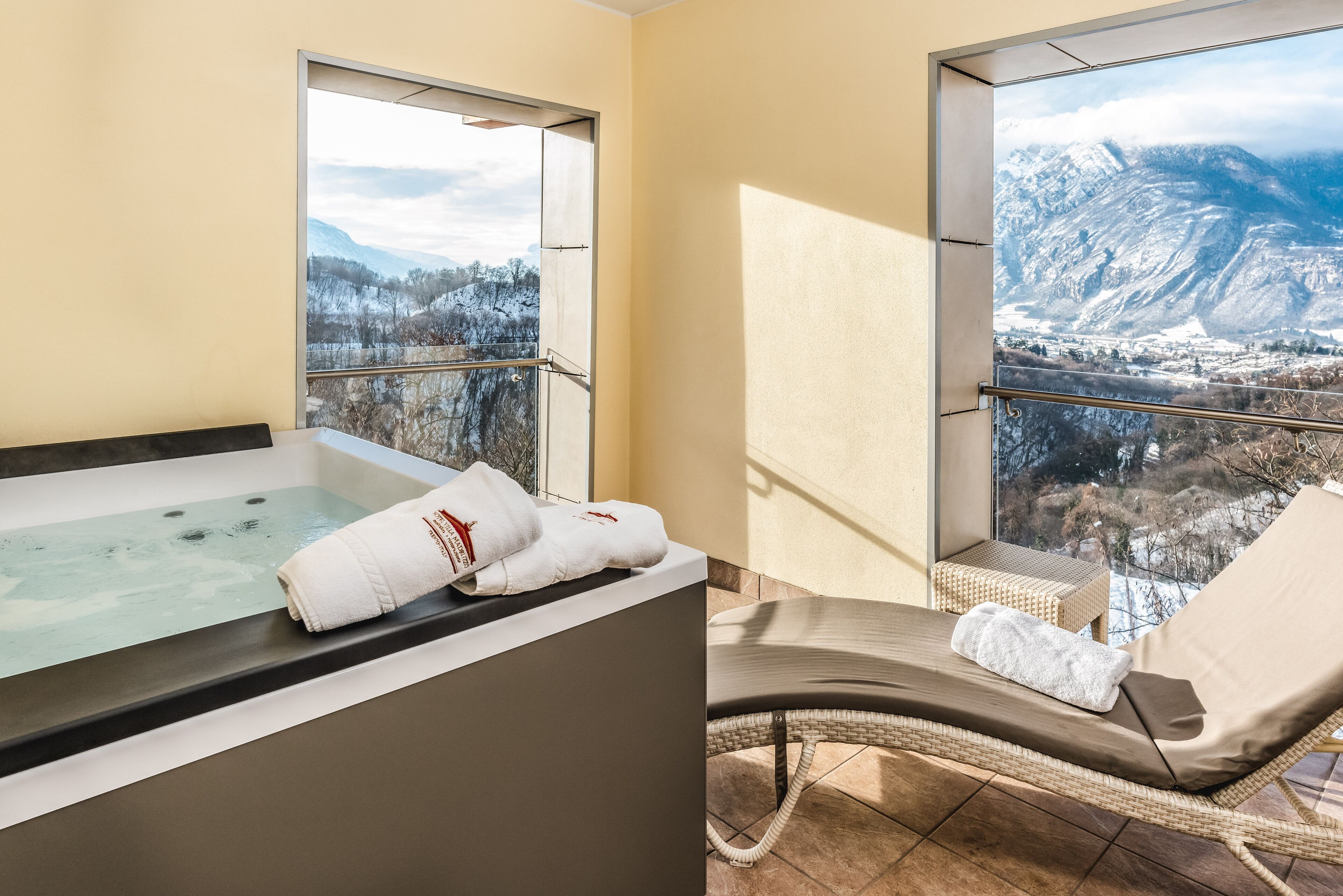 Área de spa com banheira e espreguiçadeira, ambos com toalhas, com vista para uma paisagem montanhosa coberta de neve.