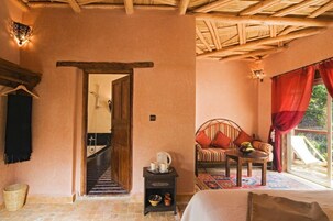 Um quarto aconchegante com uma cama, uma mesinha com xícara e pires, um cesto de vime e vista para o exterior através de uma janela com cortinas vermelhas.