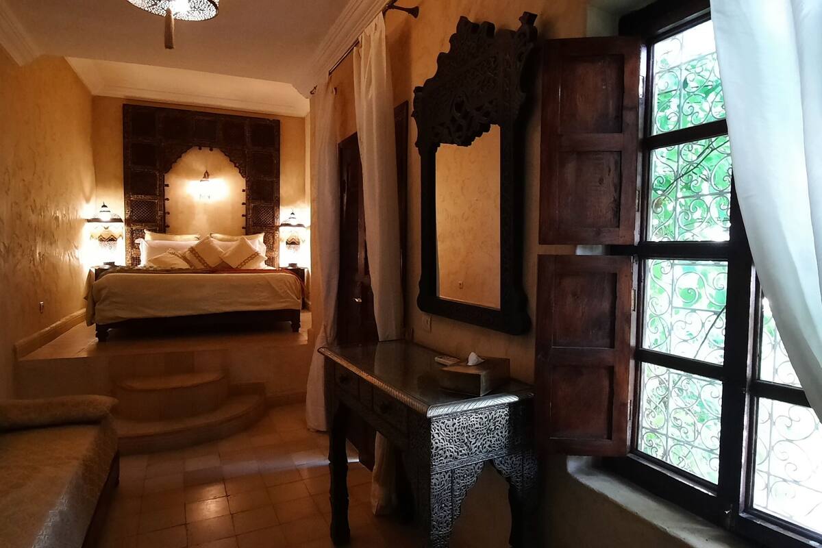 Quarto com cama, mesa de cabeceira, mesa e janela com cortinas.