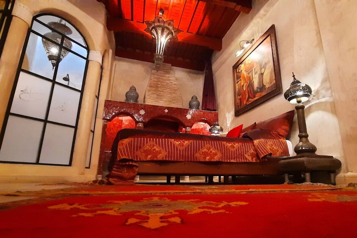 Quarto tradicional com cama, tapete vermelho, lustre e luminária na parede.