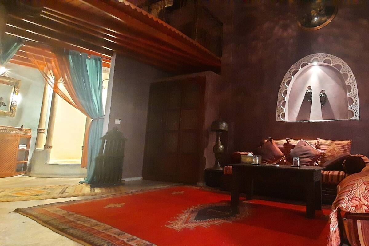 Quarto com cama, mesa e espelho. Há uma janela com moldura decorativa.