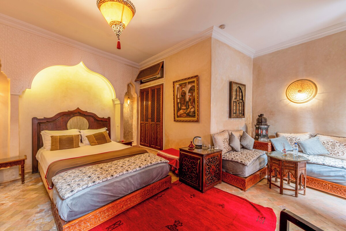 Quarto tradicional com cama grande, sofá, mesinha e detalhe decorativo na parede.