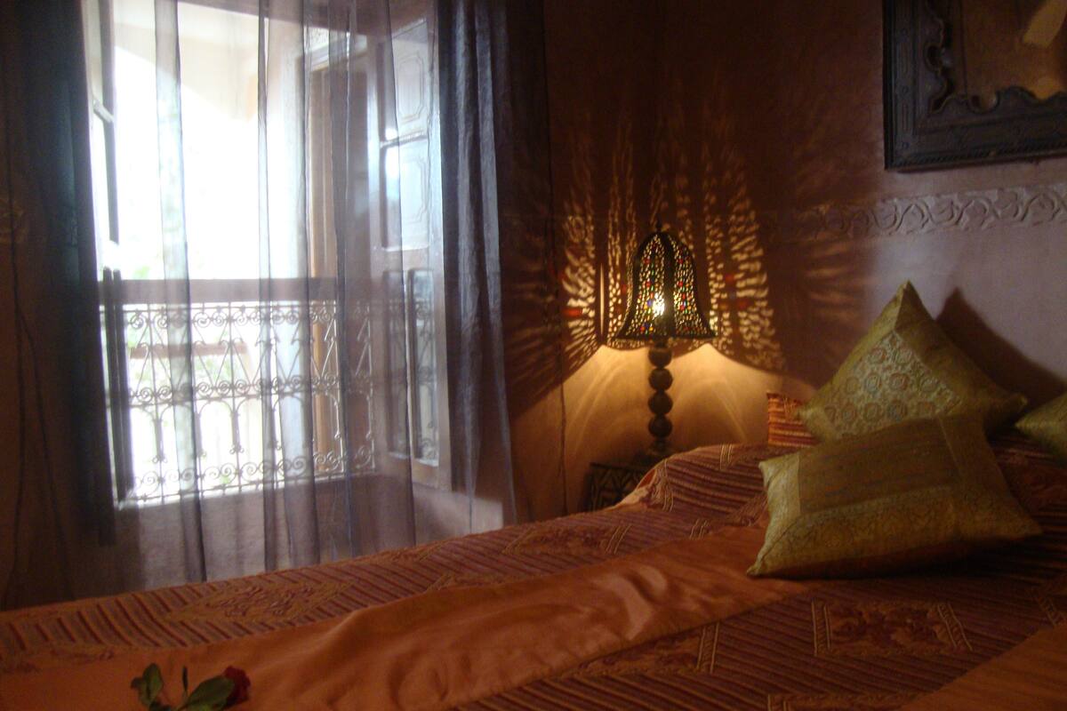Uma cama com colcha estampada, uma rosa sobre a cama, uma luminária com cúpula decorativa e uma janela com vista para o verde.