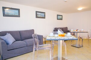 Uma sala de estar com um sofá cinzento, uma cadeira de plástico transparente, uma mesa de jantar com itens de café da manhã e quadros nas paredes.