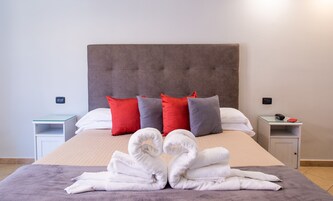 Cama bem arrumada com cabeceira cinzenta, almofadas brancas e vermelhas, e duas toalhas brancas dispostas em forma de cisne.
