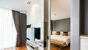 Quarto de hotel moderno com uma grande televisão de tela plana, uma cama com várias almofadas e piso de madeira.