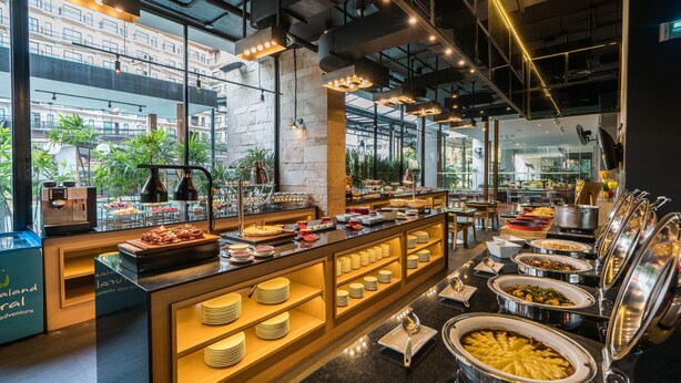 Uma área de buffet com diversos pontos de comida, uma máquina de café e vista para a paisagem urbana através de grandes janelas.